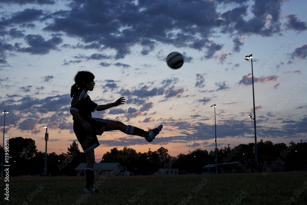 Side view of girl kicking soccer ball at field against sky during ...