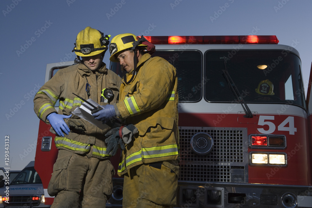 Male firefighters reading document with fire brigade in the background ...