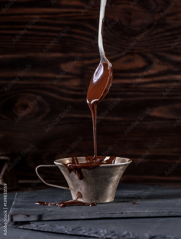Liquid chocolate dripping from spoon into cup on black stone plate and ...
