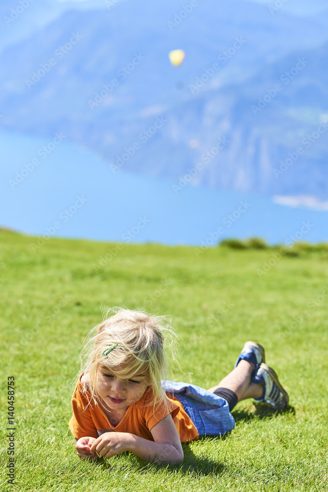 Child blond girl lying on grass over the lake Lago di Garda on the top ...