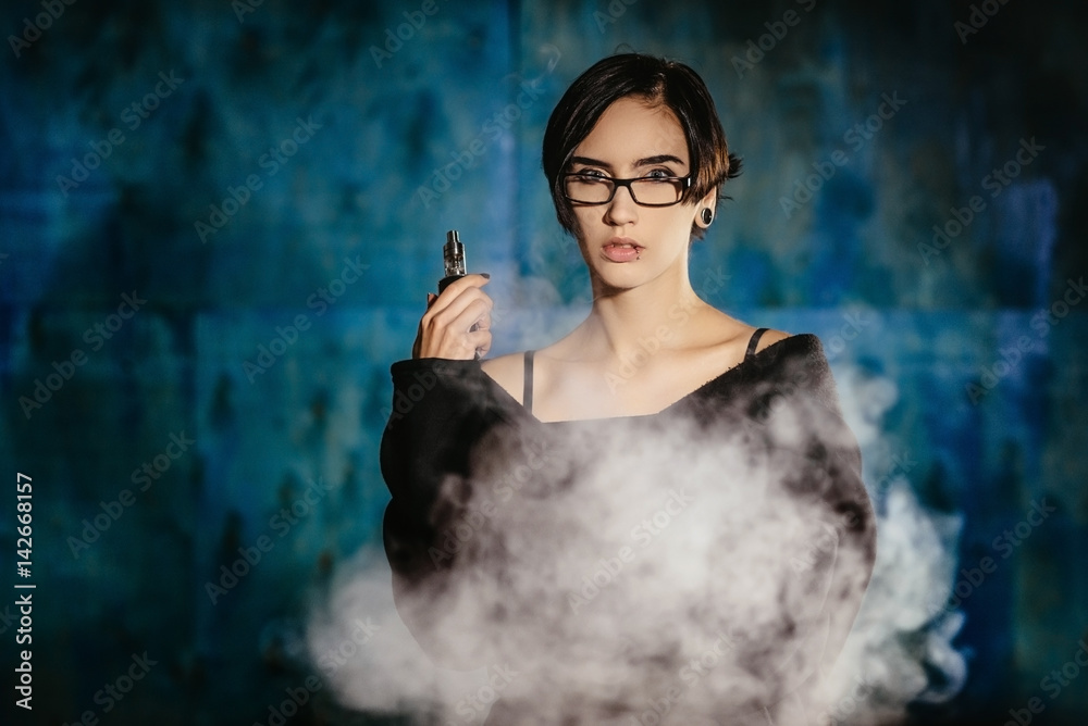 A girl with glasses vaping and releases a cloud of vapor. Model in a ...
