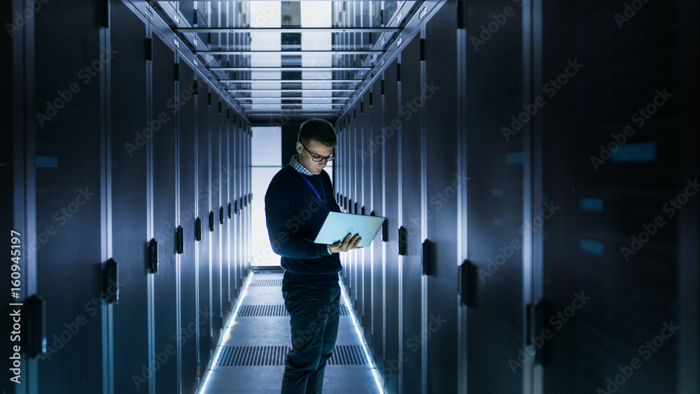 Male IT Engineer Works on a Laptop in front of Server Cabinet at a Big ...