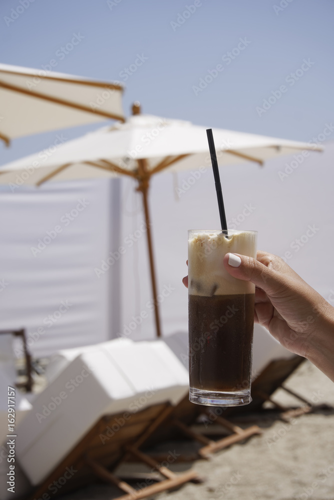 Iced coffee in hand with blurred beach background. Female hand holding ...