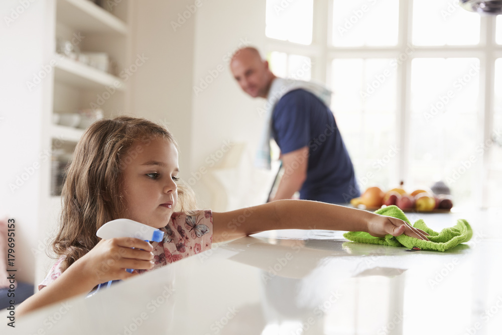 Girl cleaning kitchen worktop, father sweeping floor in the background ...