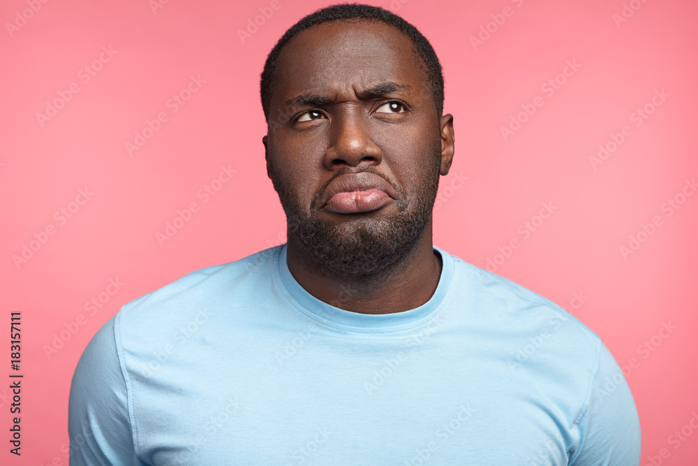 Displeased black man curves lips, being deep in thoughts, looks upwards ...