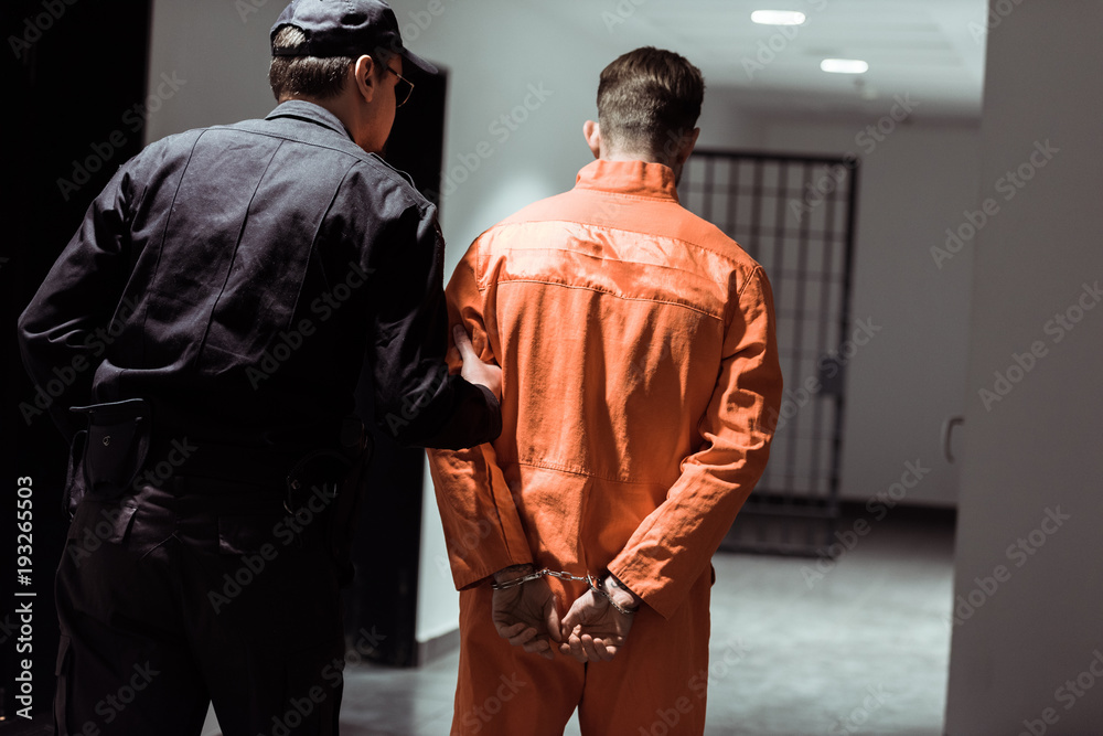 rear view of prison officer leading prisoner in handcuffs in corridor ...