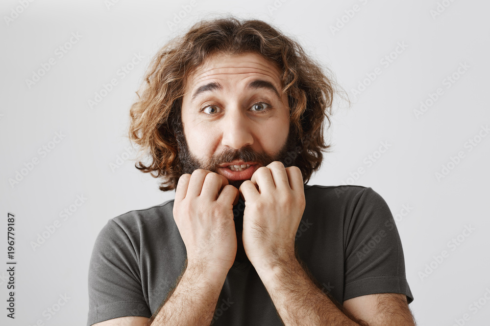 Man trembling from fear. Indoor shot of scared adult bearded guy with curly hair holding hands ...
