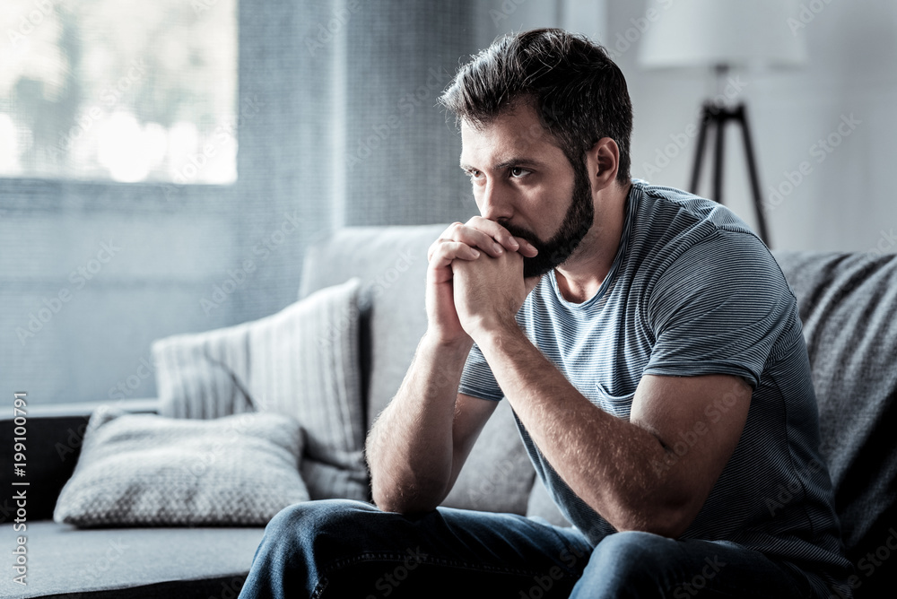 Alone at home. Serious nice thoughtful man sitting on the bed and ...