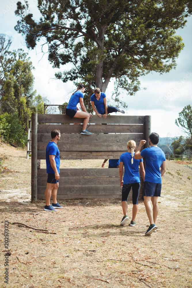 Female trainer assisting fit woman to climb over wooden wall during ...