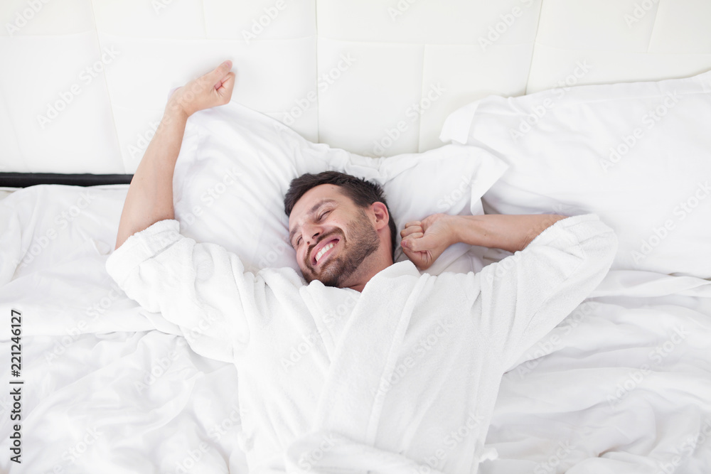 Cheerful young man is waking up after sleeping in the morning. He is yawing and stretching his ...