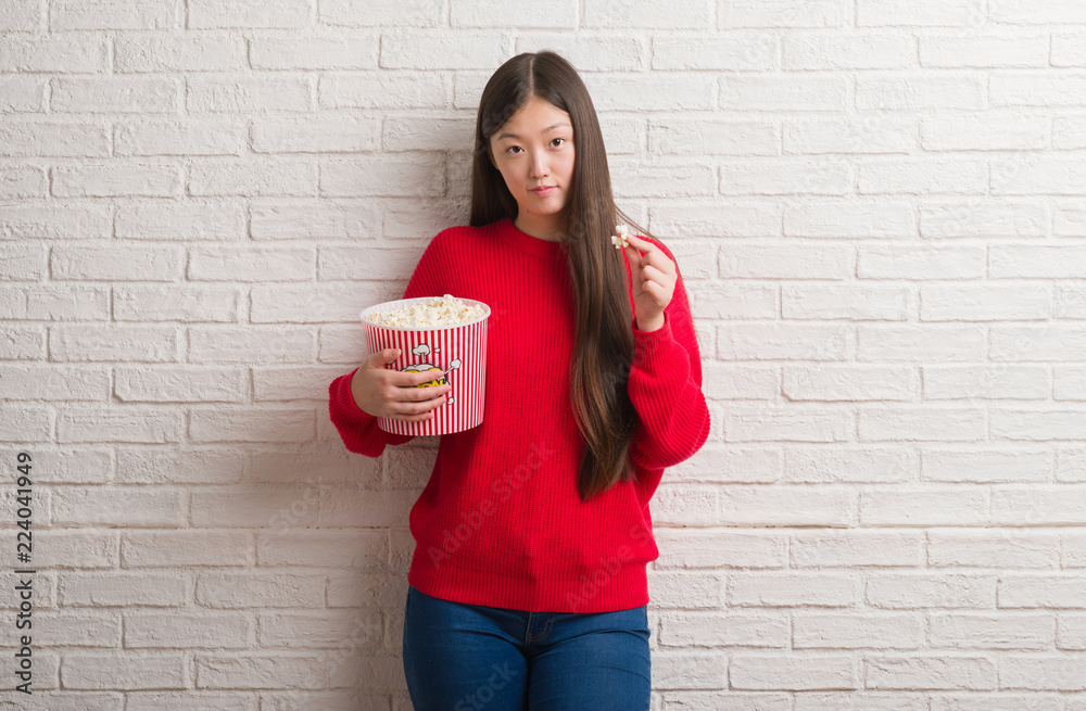 Young Chinese woman over brick wall eating pop corn with a confident ...