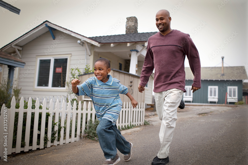 Smiling mid-adult father running down a suburban street with his young ...