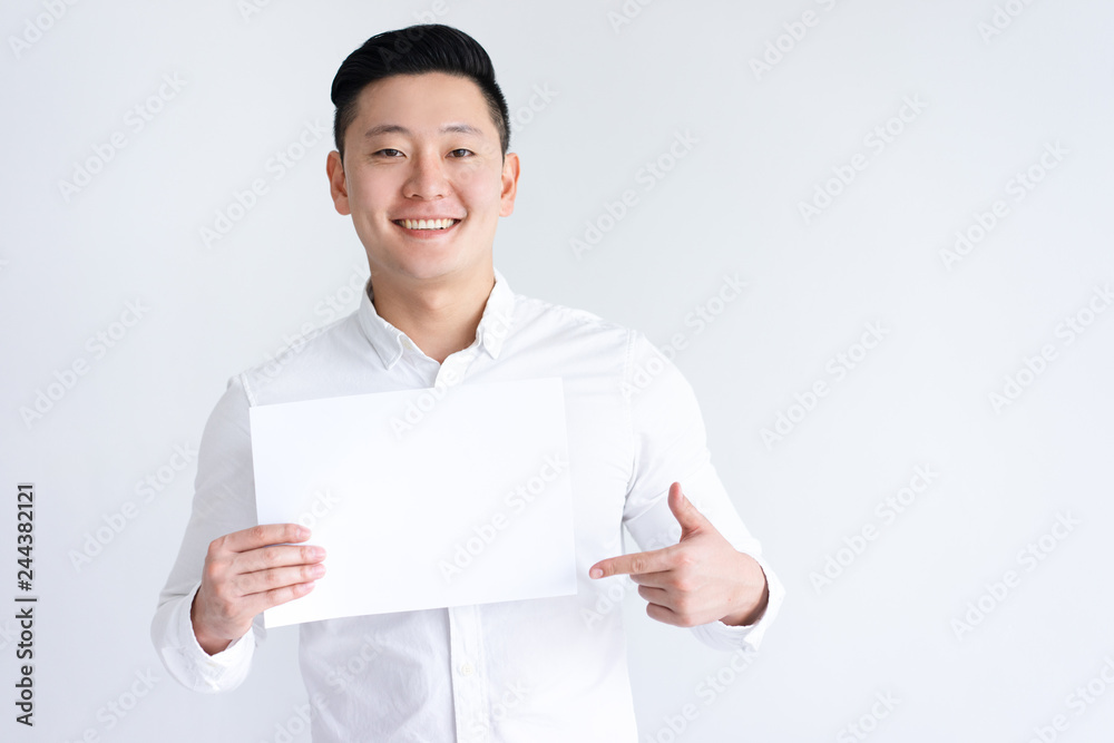 Smiling Asian man holding blank sheet of paper. Young guy pointing at ...