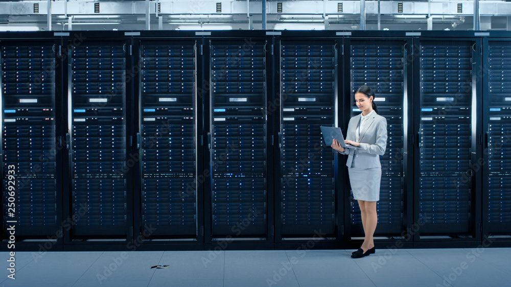 Female Data Center IT Engineer Works on Laptop Computer Next to Server Rack Corridor. Running ...