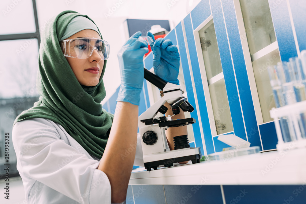 female muslim scientist sitting at table with microscope and looking at ...