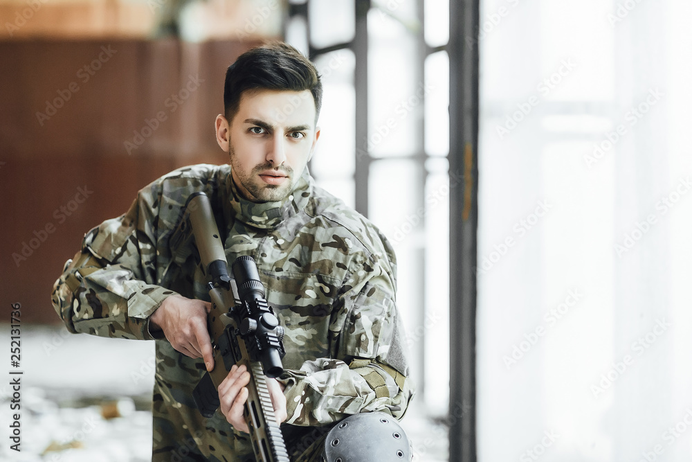 A young military soldier sits with a big rifle in his hands, near the ...