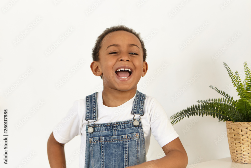 Cute emotional dark skinned little boy posing at white wall with plant ...