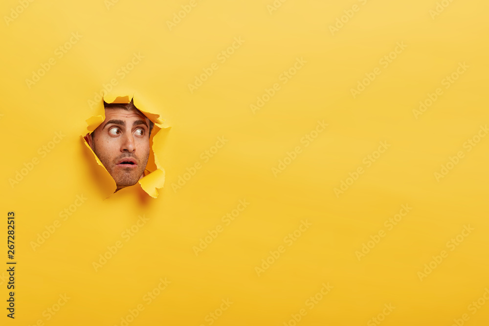 Astonished unshaven man has stupefied expression, peeks through torn ...