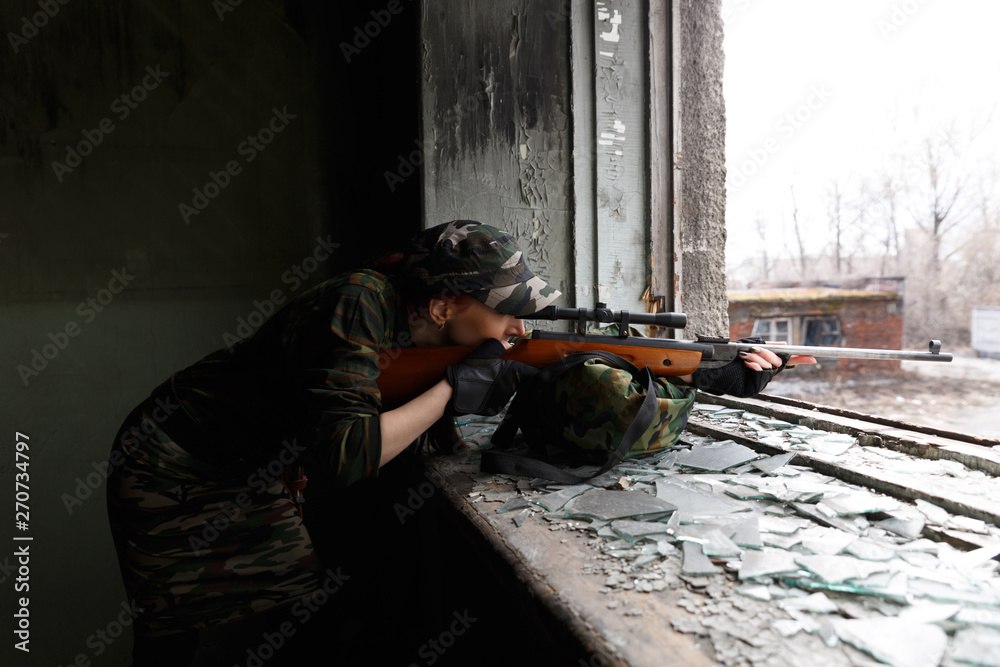 Girl sniper looks through the scope. Girl warrior directs his rifle through the broken window of ...
