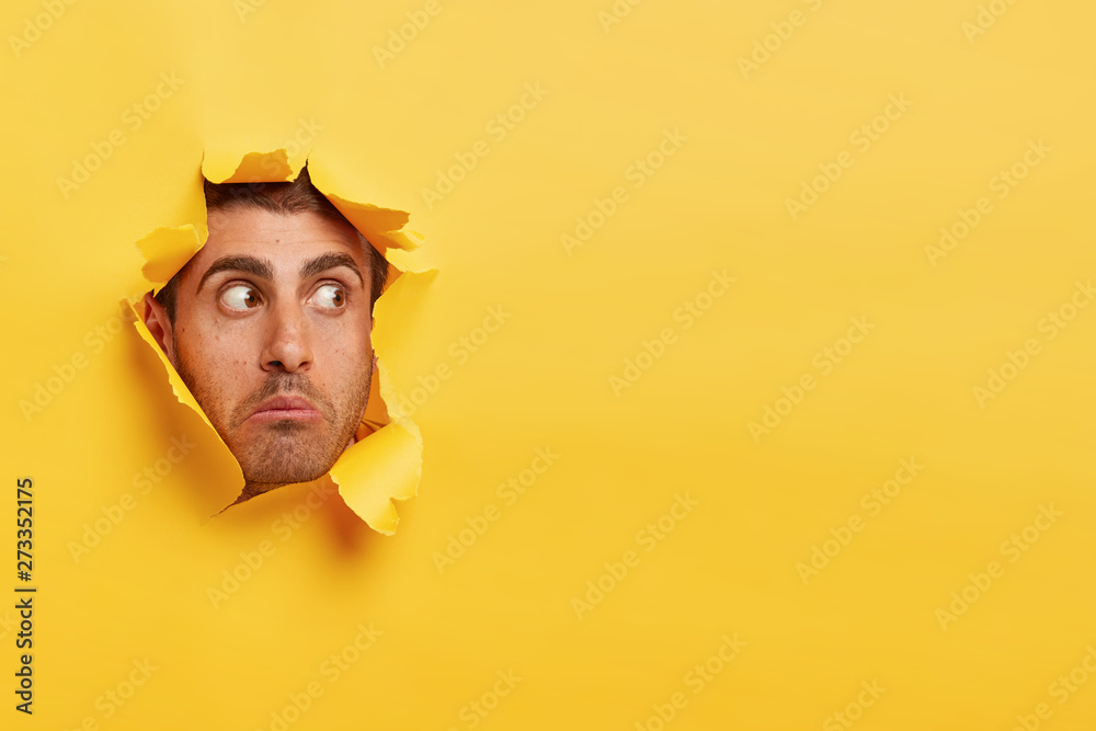 Face of surprised scared young man gazes through yellow paper hole ...