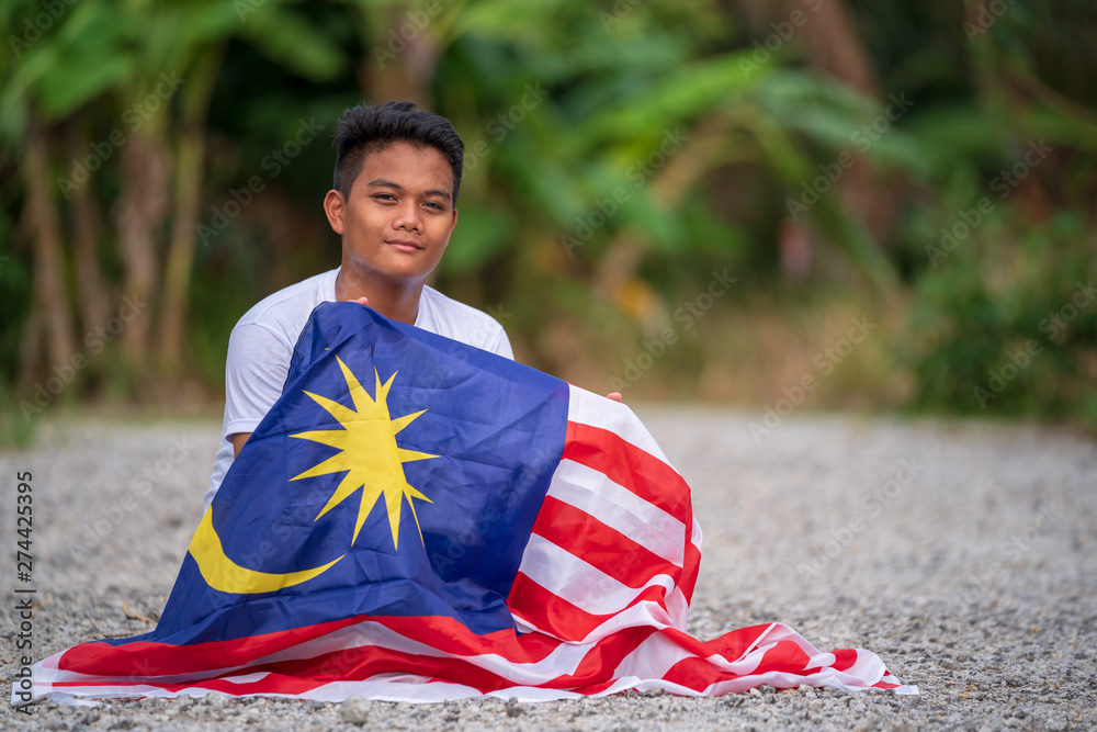 Portrait of young Malay Asian man sitting and holding Malaysian flag or ...