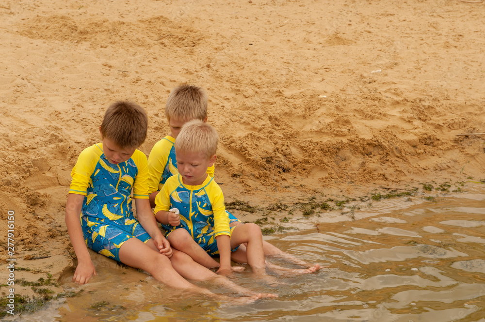 Funny kids in bathing suits sit together on sand with their legs in ...