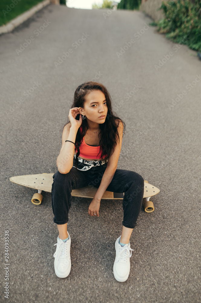 Beautiful girl sits on a skateboard and looks into the camera, wears ...