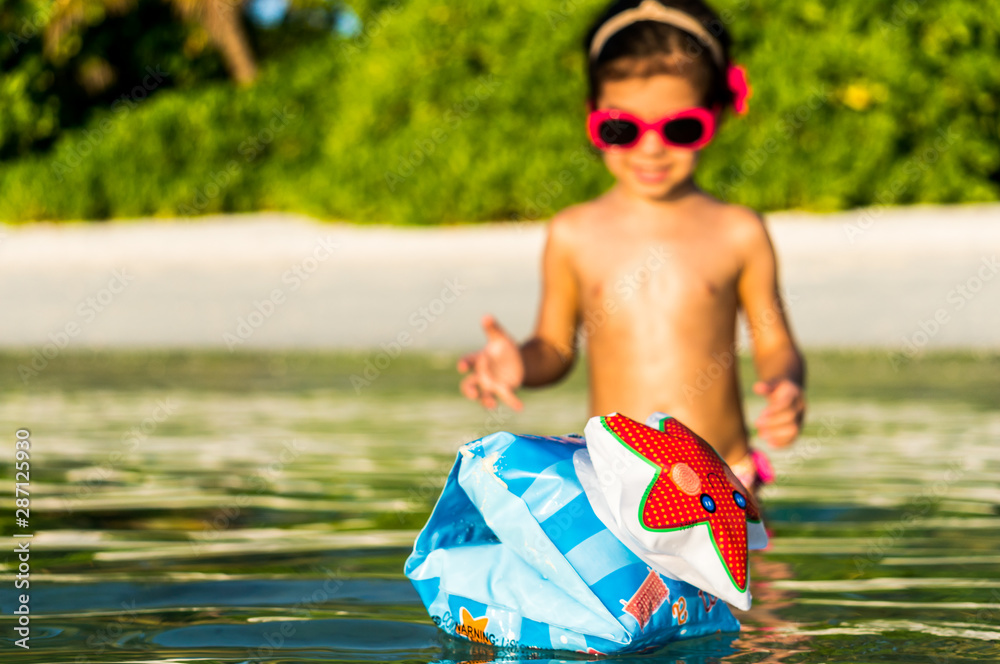 Little girl in water on beach with tropical flora and inflatable arm ...