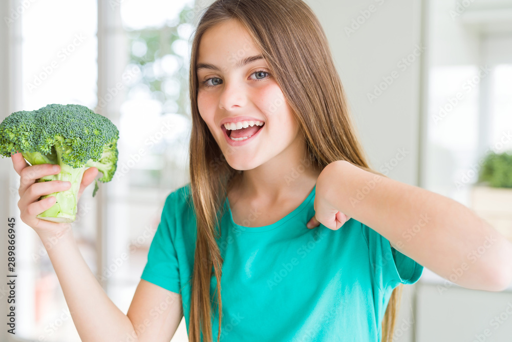Beautiful young girl eating fresh broccoli with surprise face pointing ...