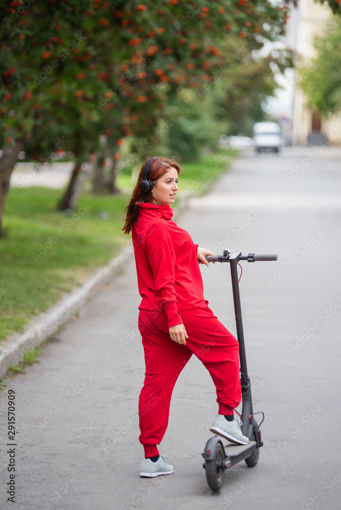 Red-haired girl in a red tracksuit drives an electric scooter. A young ...