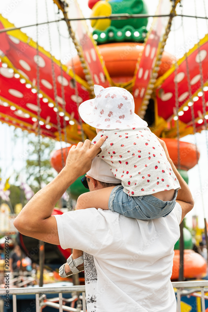 Child sitting on her father's shoulders in front of a colorful carousel ...