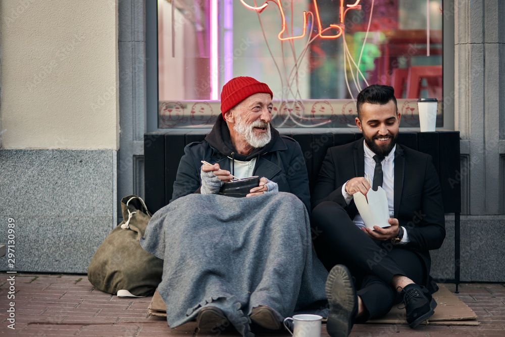 Smiling rich and poor men together sitting on street and eat while ...
