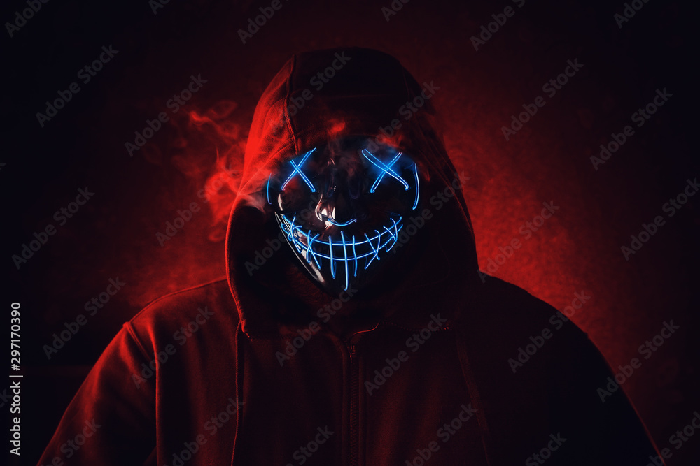 Man in angry and scary lighting neon glow mask in hood on dark red ...