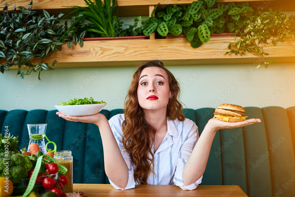 Beautiful young woman decides eating hamburger or fresh salad in ...