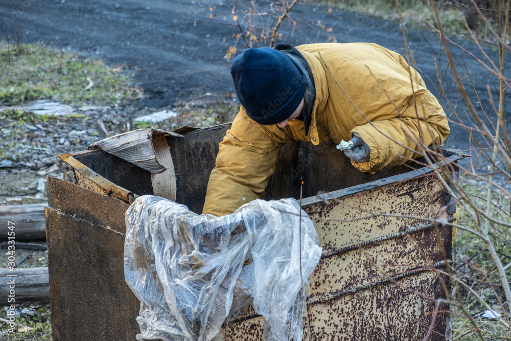 homeless woman in a yellow old ragged jacket and blue hat is eating ...