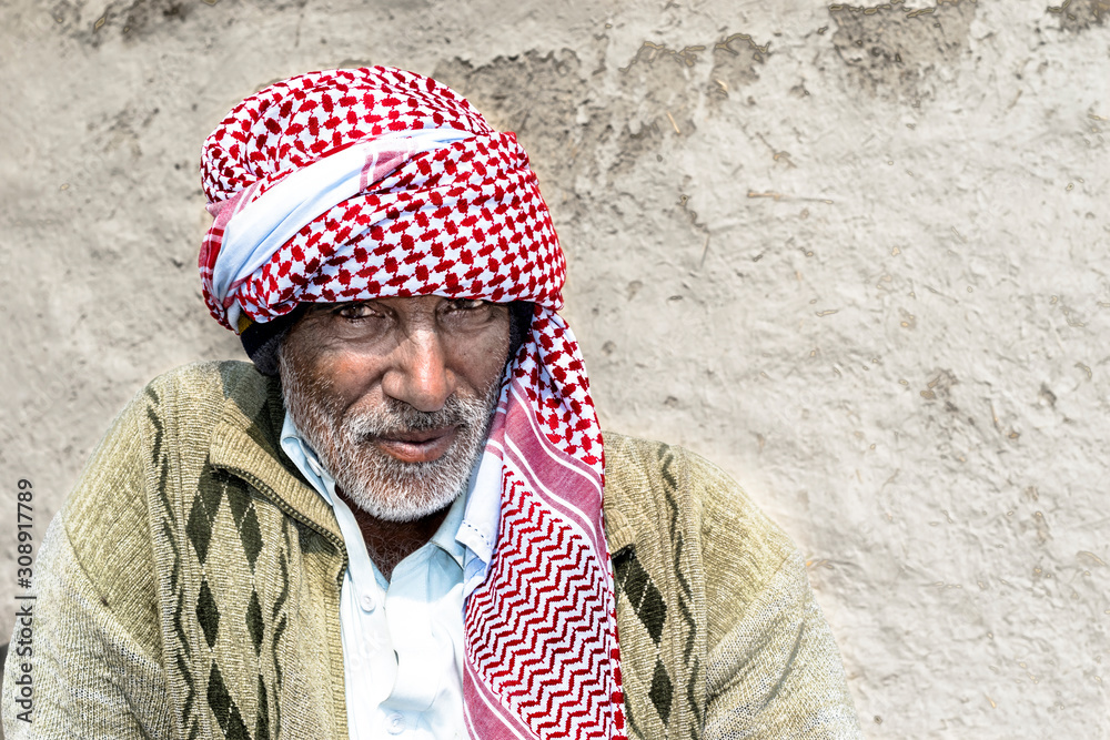 a homeless an old Arabic man is sitting under sunshine and wearing ...