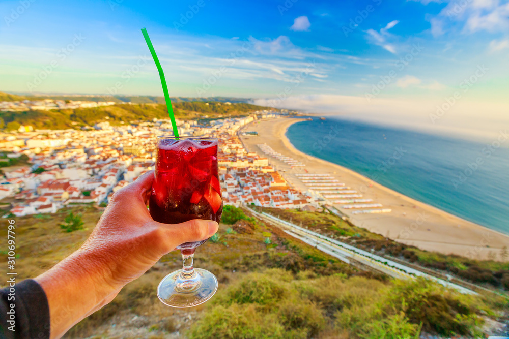 Glass of Sangria in man hand on Nazare skyline blurred background ...