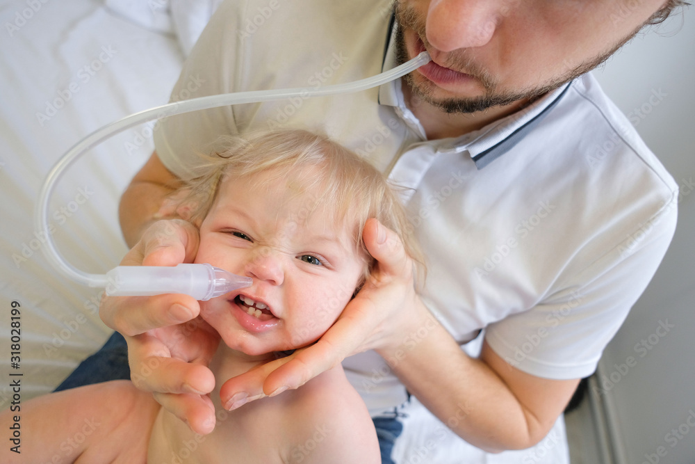 in the white room, a baby with snot sits on dad’s lap. Dad and son are ...