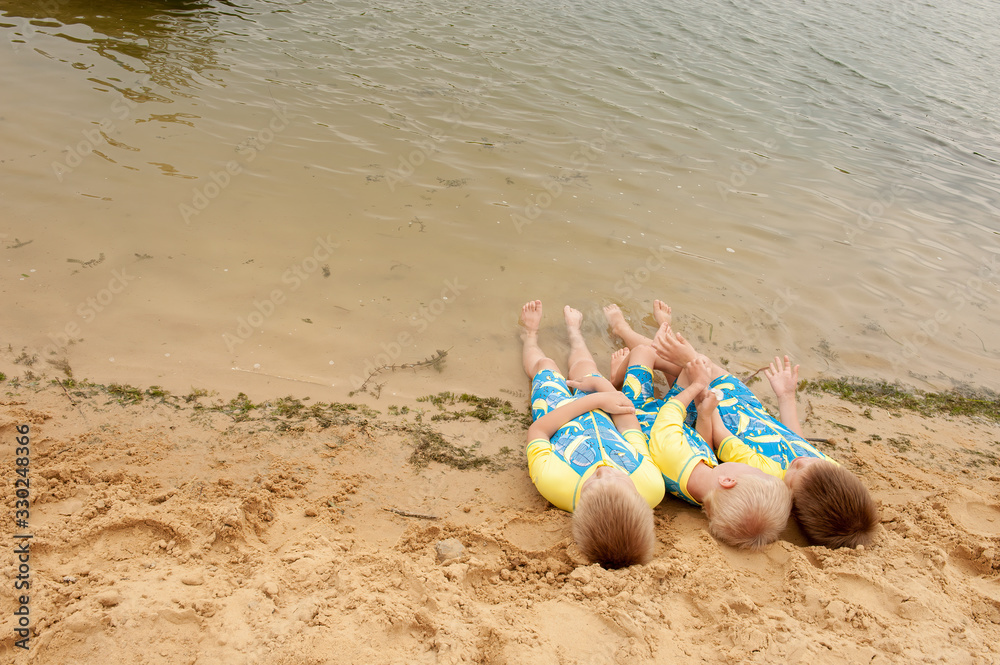 Funny kids in bathing suits sit together on sand with their legs in ...