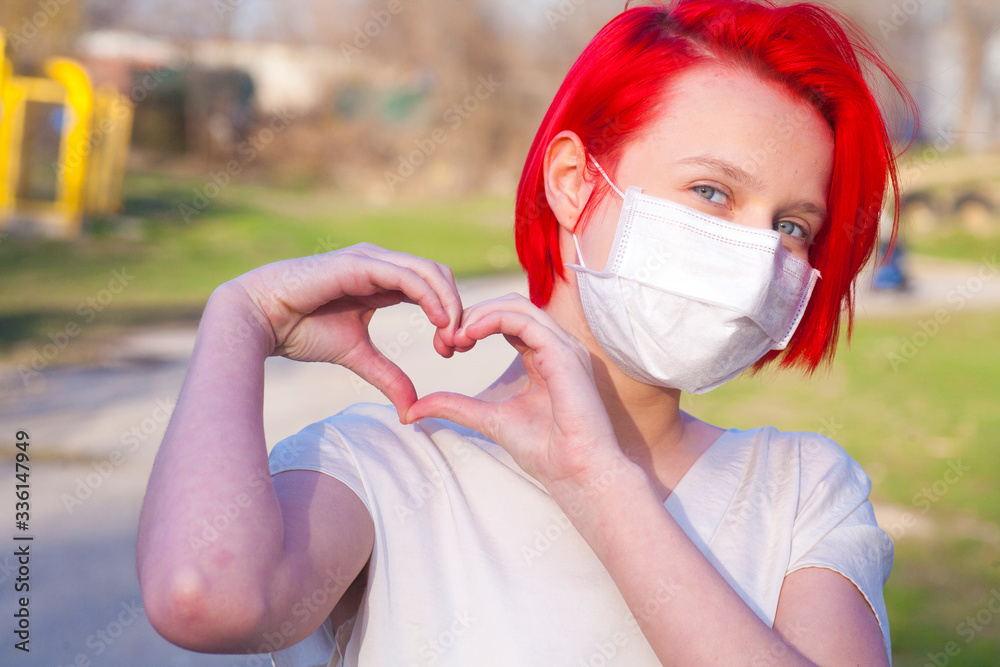 Red hair teenage girl wearing a medical mask shows the heart symbol ...