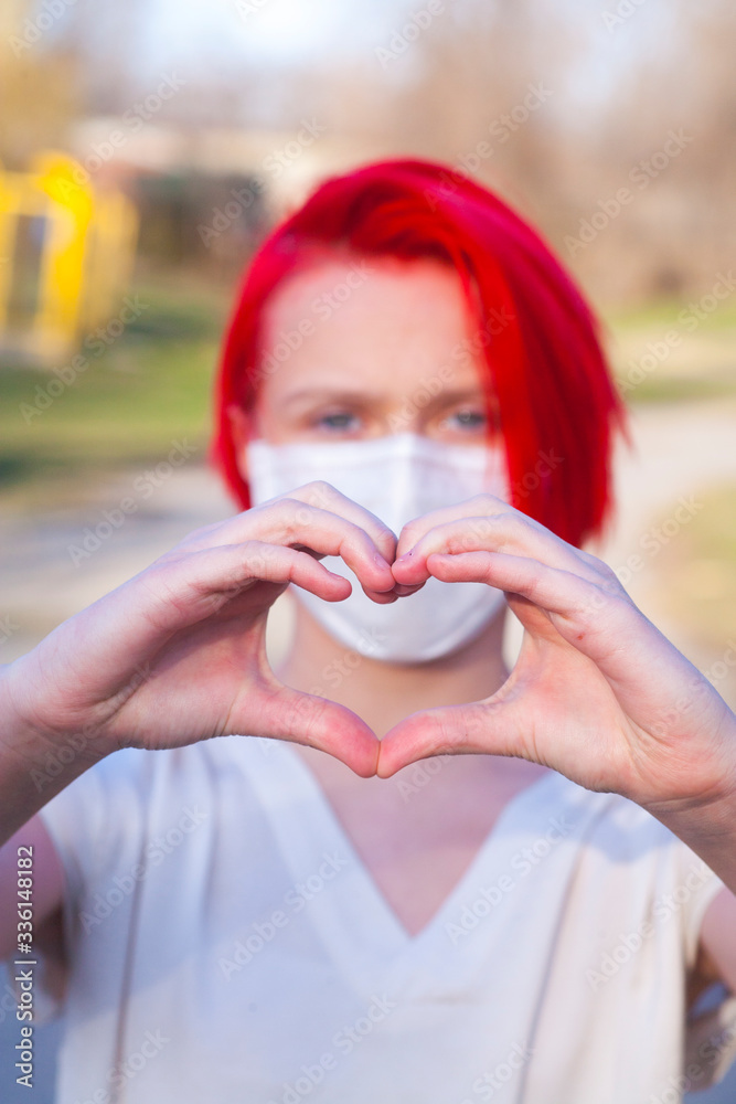 Red hair teenage girl wearing a medical mask shows the heart symbol ...