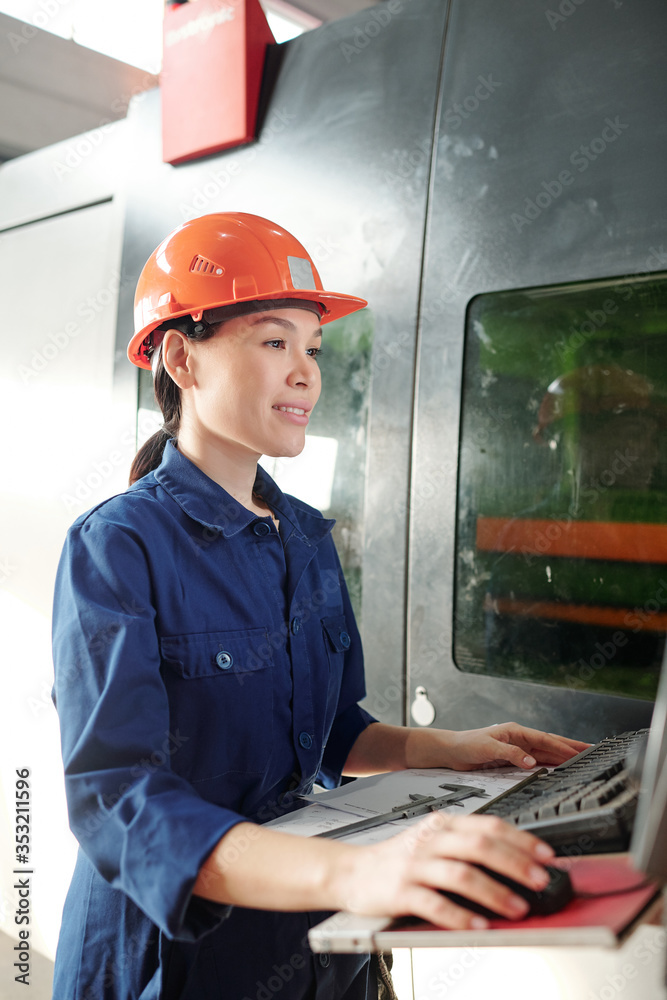 Smiling young Asian woman in hardhat operating CNC machine while ...