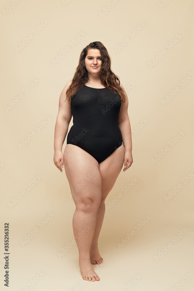 Plus Size Model. Big Woman In Black Bodysuit Full Length Portrait. Brunette Standing And Looking At Camera. Body Positive Concept On Beige Background. Stock Photo Adobe Stock