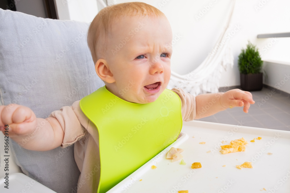 Annoyed cute baby wearing plastic bib, sitting in highchair with food messy on tray. First solid ...