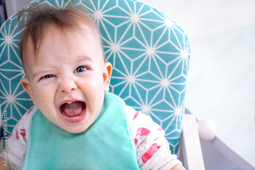 Positive baby having lunch in a bib, sitting in highchair. First solid food or child care at ...