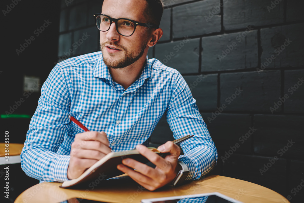 Handsome thoughtful young man in stylish eyewear writing down notes in ...