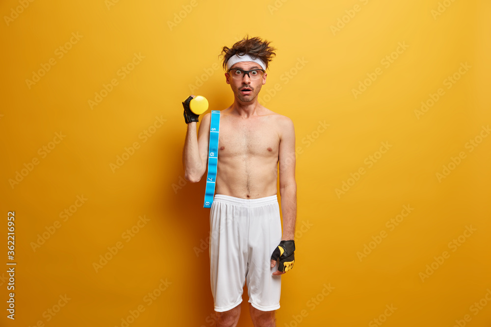 Studio shot of shocked sporty man holds dumbbell, tape measure on ...