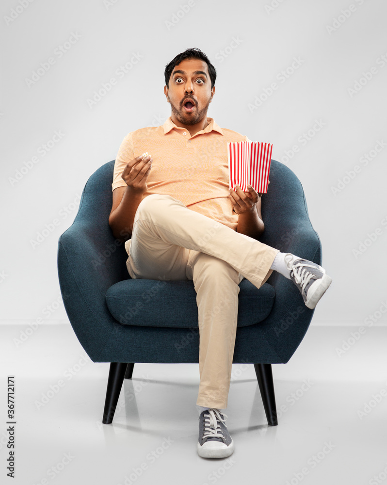 people and furniture concept - shocked young indian man eating popcorn ...