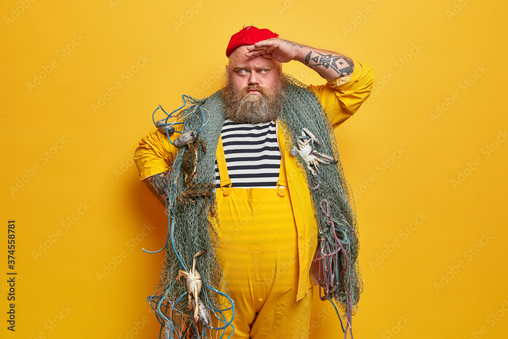 Bearded serious fisher keeps hand on forehead and looks into distance ...