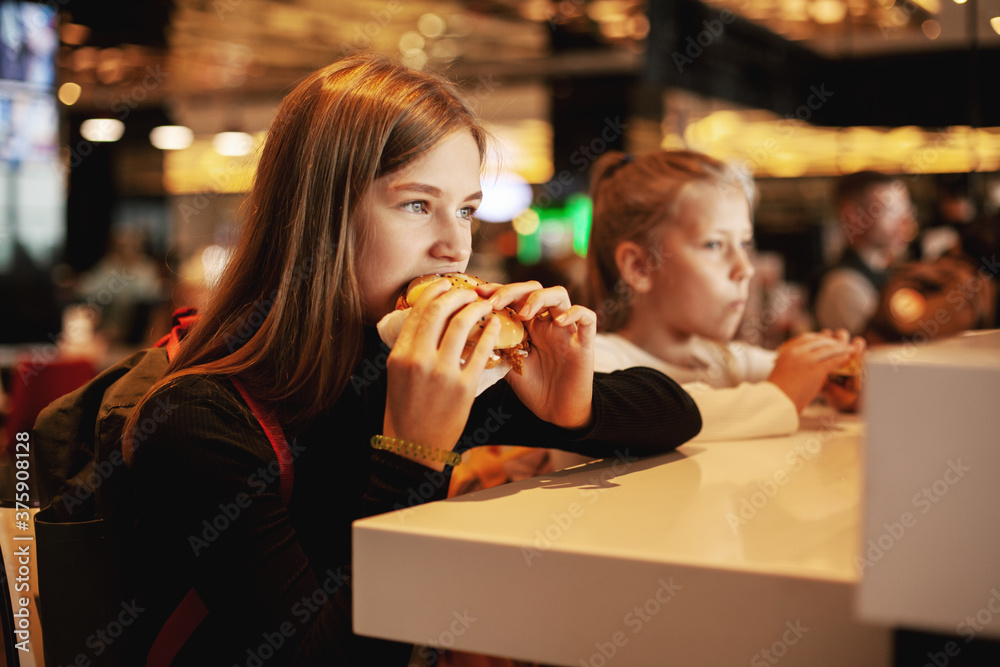 Beautiful schoolgirl eating hamburger sitting in a cafe indoors. Happy ...